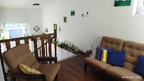 Uma sala de estar com escada de madeira, sofá e uma cadeira. Há várias plantas em vasos na escada e uma janela com vista para o exterior.