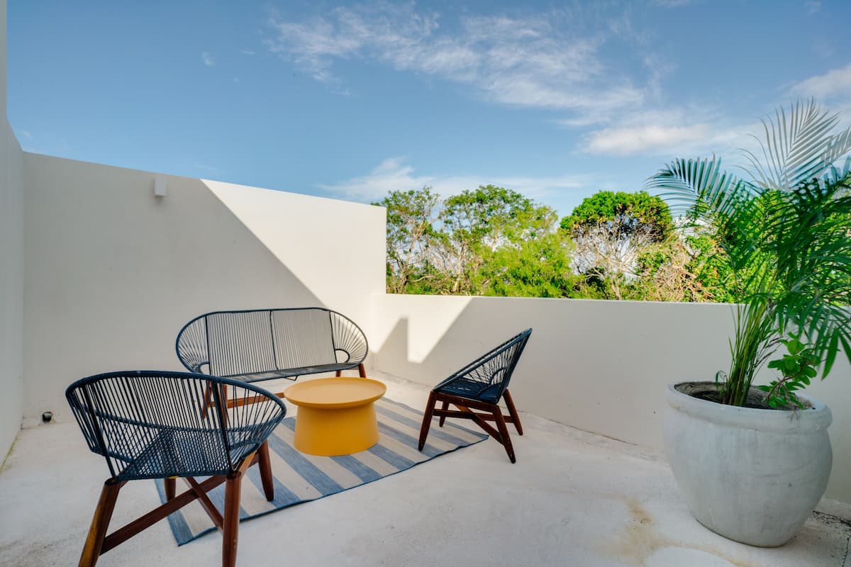 Uma área externa com assentos de metal, cadeiras, uma mesa redonda, um vaso de planta e vista para o verde.