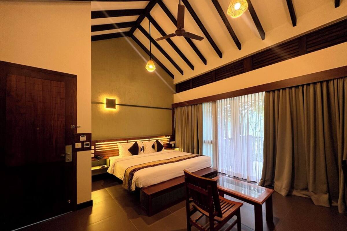 Quarto de hotel com cama grande, escrivaninha, cadeira, ventoinha no teto e janela com cortinas.