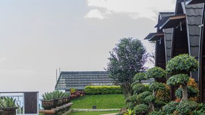 Um caminho leva a um edifício com telhado inclinado, cercado por vegetação bem cuidada e vasos de plantas.