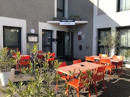 Uma área externa com mesas e cadeiras, uma placa indicando um hotel e café, e uma entrada de prédio com porta e janelas.