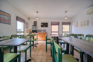 Um restaurante com cadeiras verdes e mesas de madeira, uma área de cozinha e uma televisão na parede.
