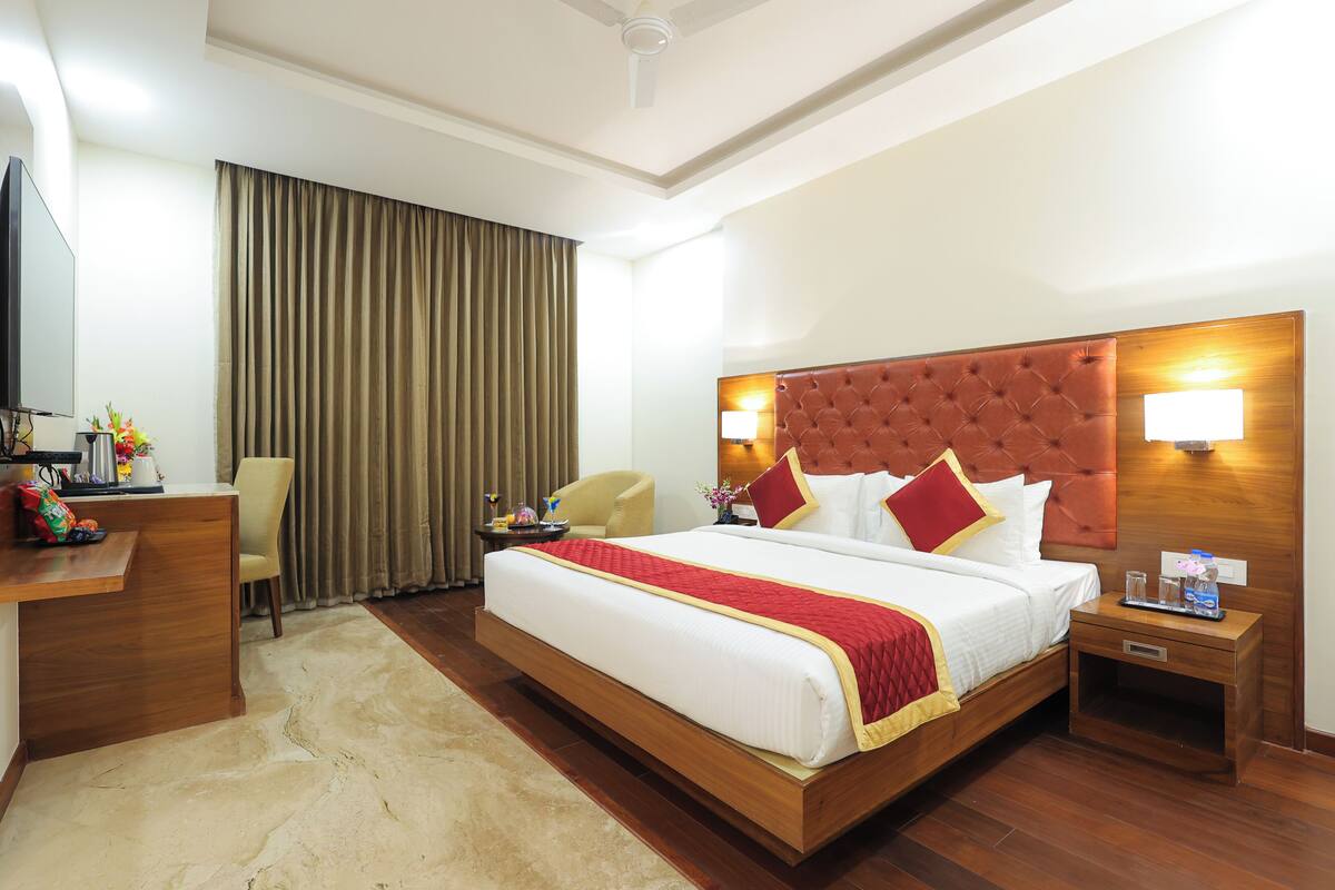 Quarto de hotel com uma cama grande, uma escrivaninha, uma cadeira, uma televisão e uma janela com cortinas.
