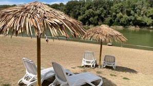 Uma praia com cadeiras de praia brancas e guarda-sóis de palha, um lago calmo e uma área arborizada ao fundo.
