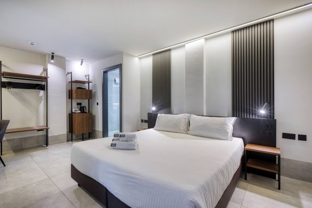 Quarto de hotel moderno com uma cama grande, um banco de madeira, um guarda-roupa e uma pequena mesa com uma chaleira.