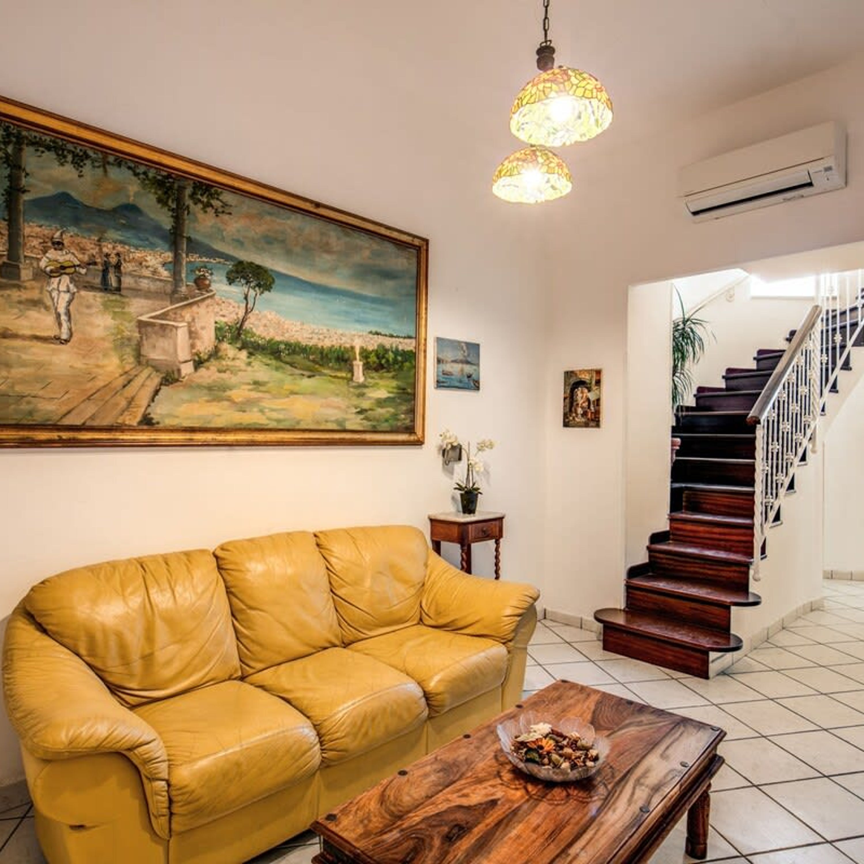 Uma sala de estar com um sofá de couro amarelo, uma mesa de centro de madeira com uma tigela, um quadro na parede, uma escada e um aparelho de ar condicionado.