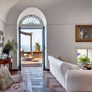 Quarto com sofá branco, mesa de centro e vista através de uma porta em arco para um pátio externo com mesa e cadeiras.