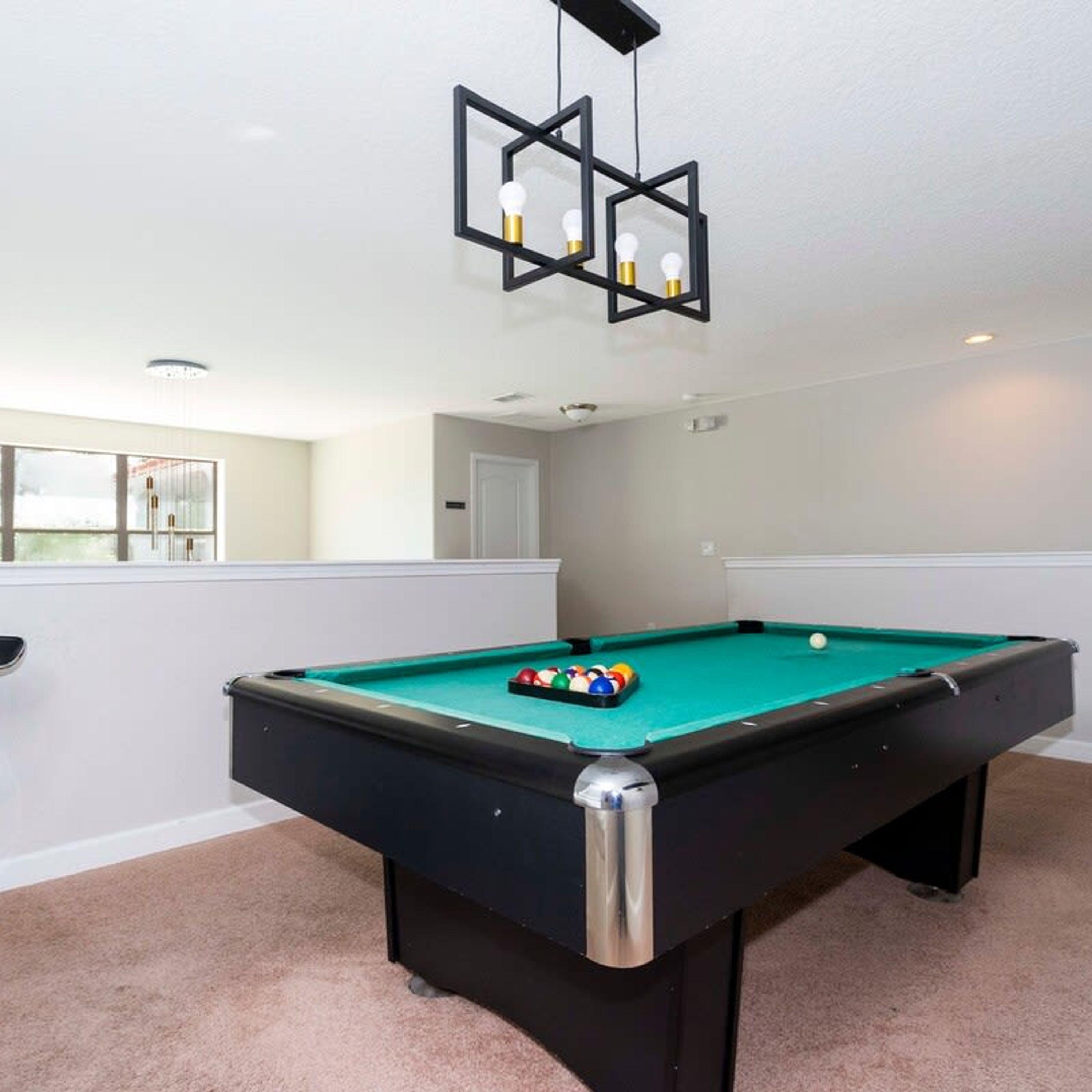 Uma mesa de bilhar com bolas, um candeeiro pendente e uma porta num espaço com paredes brancas e carpete.