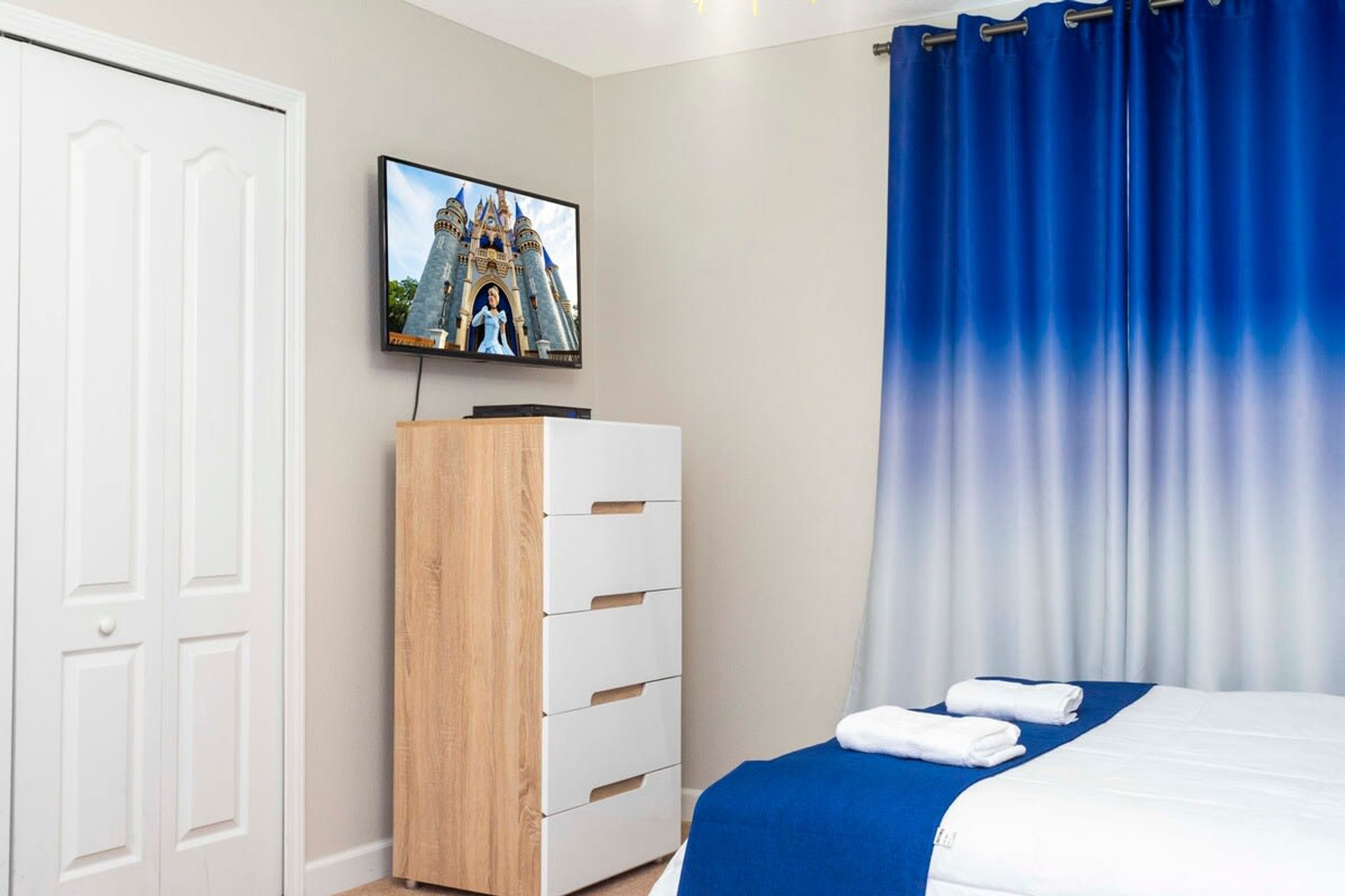 Quarto de hotel com cama, televisão presa na parede, cómoda de madeira e cortina azul.