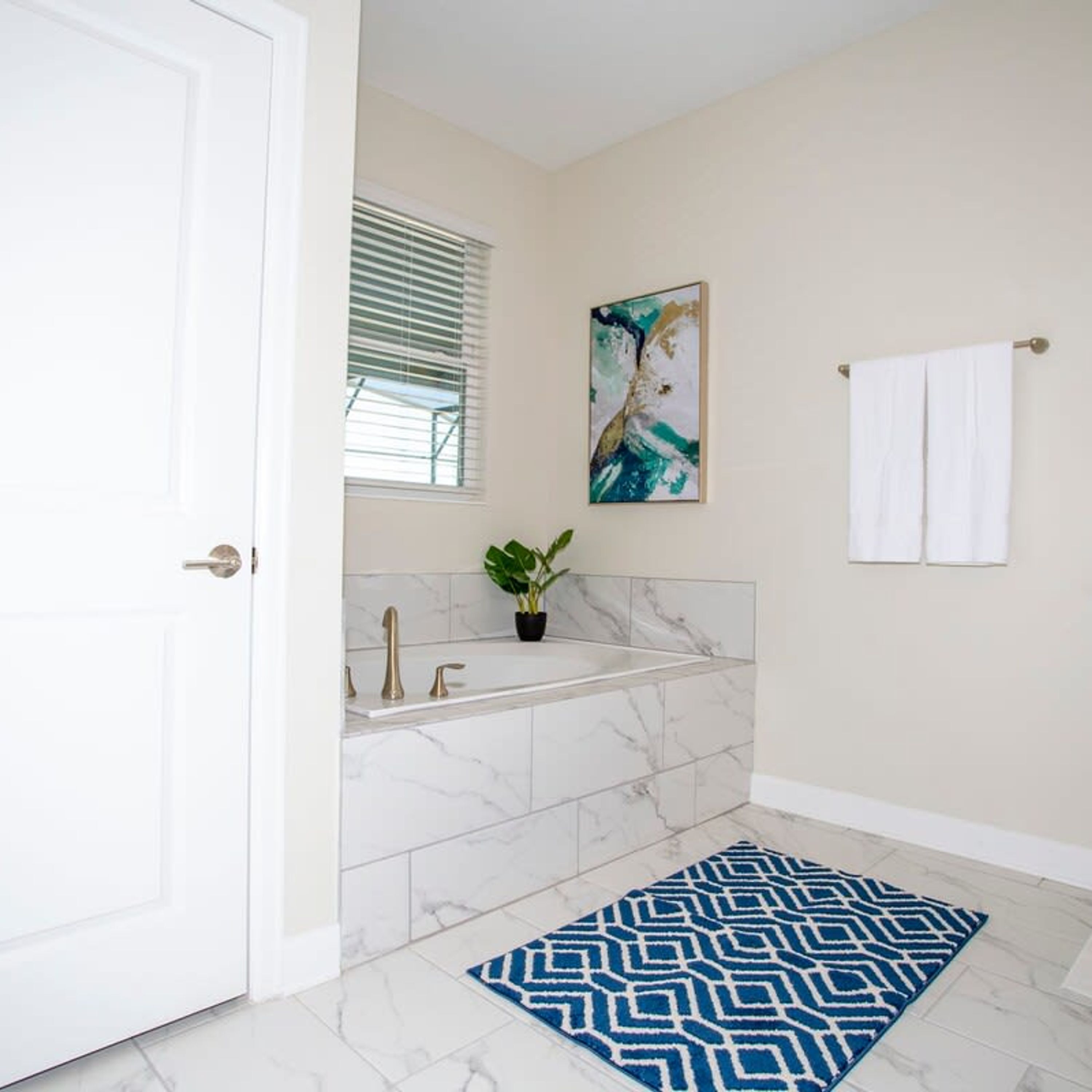 Um banheiro com porta branca, pia de mármore, planta em vaso, tapete azul estampado e um quadro na parede.