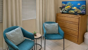 Quarto com dois poltronas cor turquesa, uma mesinha redonda com uma planta, um armário de madeira e uma televisão exibindo uma cena subaquática.