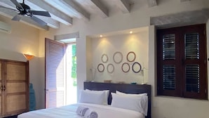 Quarto com cama de madeira, ventoinha no teto, janela com persianas e decoração circular fixada na parede.