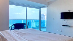 Quarto de hotel com uma cama grande, televisão na parede e varanda com vista para o mar e edifícios.