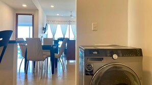 Uma lavanderia com máquina de lavar e secar de abertura frontal, uma área de refeições com mesa e cadeiras, e uma janela com cortinas.
