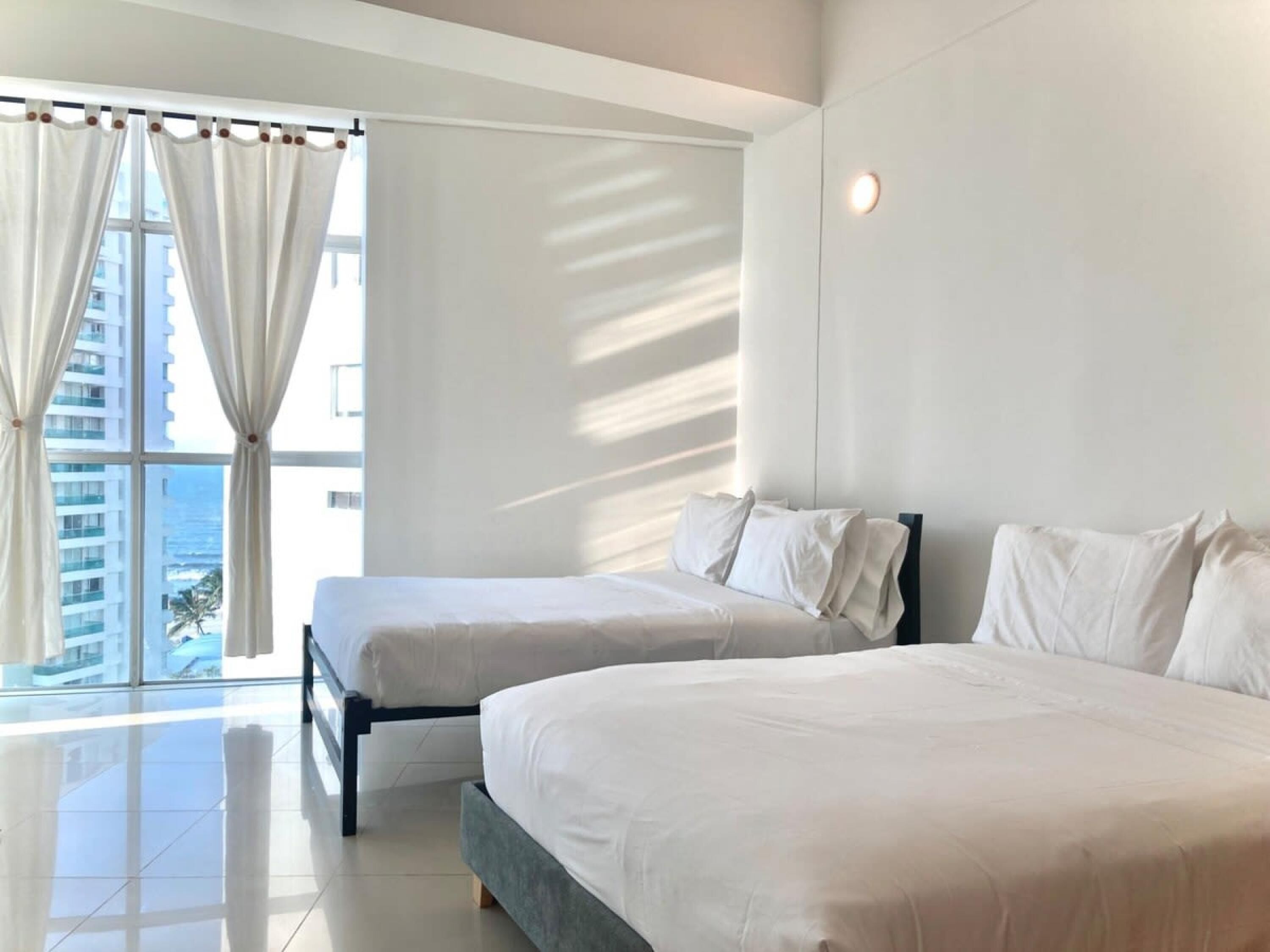 Quarto moderno com duas camas, janelas amplas com cortinas brancas e vista para edifícios e o oceano.