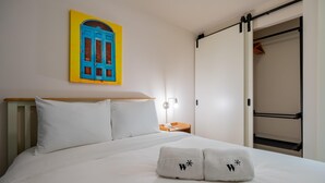 Uma cama bem arrumada com lençóis brancos e duas toalhas com logotipo, uma mesa de cabeceira com abajur, um quadro na parede de uma porta azul e uma porta de guarda-roupa deslizante.