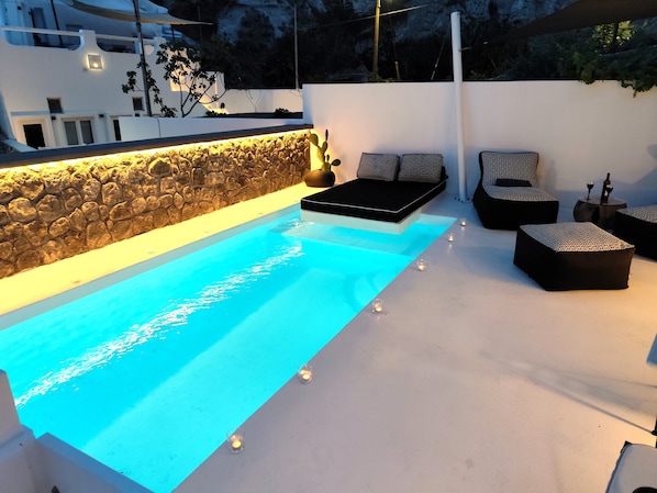 Uma área moderna com piscina ao ar livre, parede de pedra, assentos de lounge e uma pequena mesa com velas.