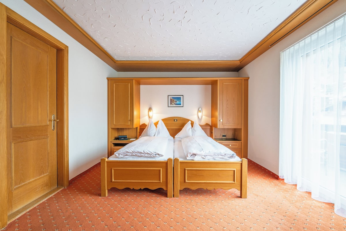 Quarto de hotel com cama de casal, guarda-roupa de madeira e pequena mesa de trabalho com abajur.