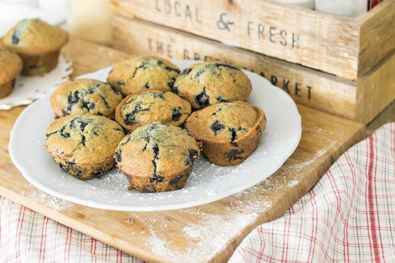 Um prato de muffins de blueberry sobre um prato branco, com uma caixa de madeira com a etiqueta 'LOCAL & FRESH' ao fundo.