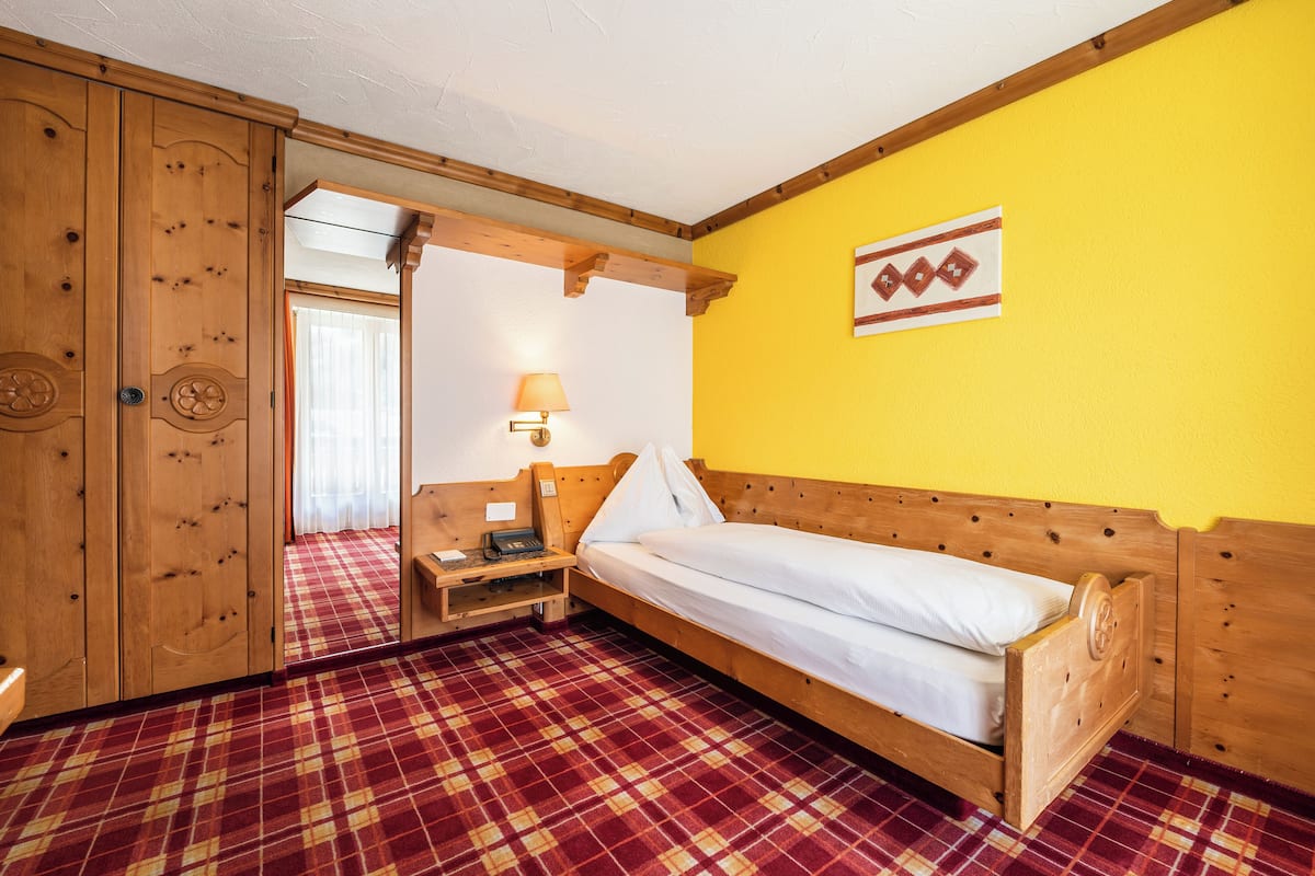 Quarto com cama de madeira, tapete xadrez vermelho, guarda-roupa de madeira e parede amarela.