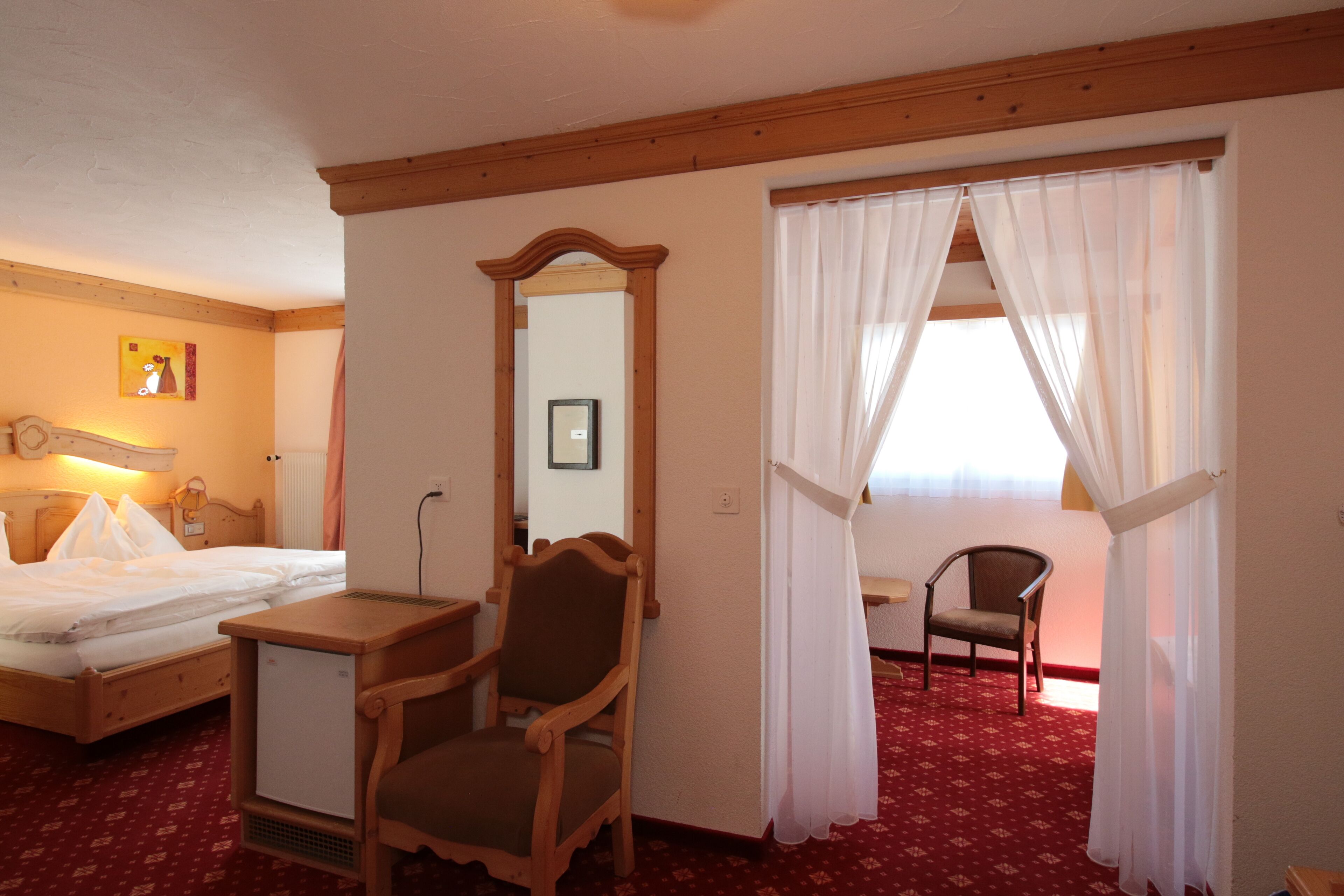 Quarto de hotel com duas camas, uma cadeira de madeira, uma escrivaninha, um espelho e uma janela com cortinas brancas.