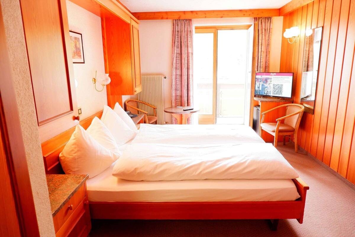 Quarto de hotel com uma cama grande, uma mesa com cadeira, televisão e vista para o exterior.