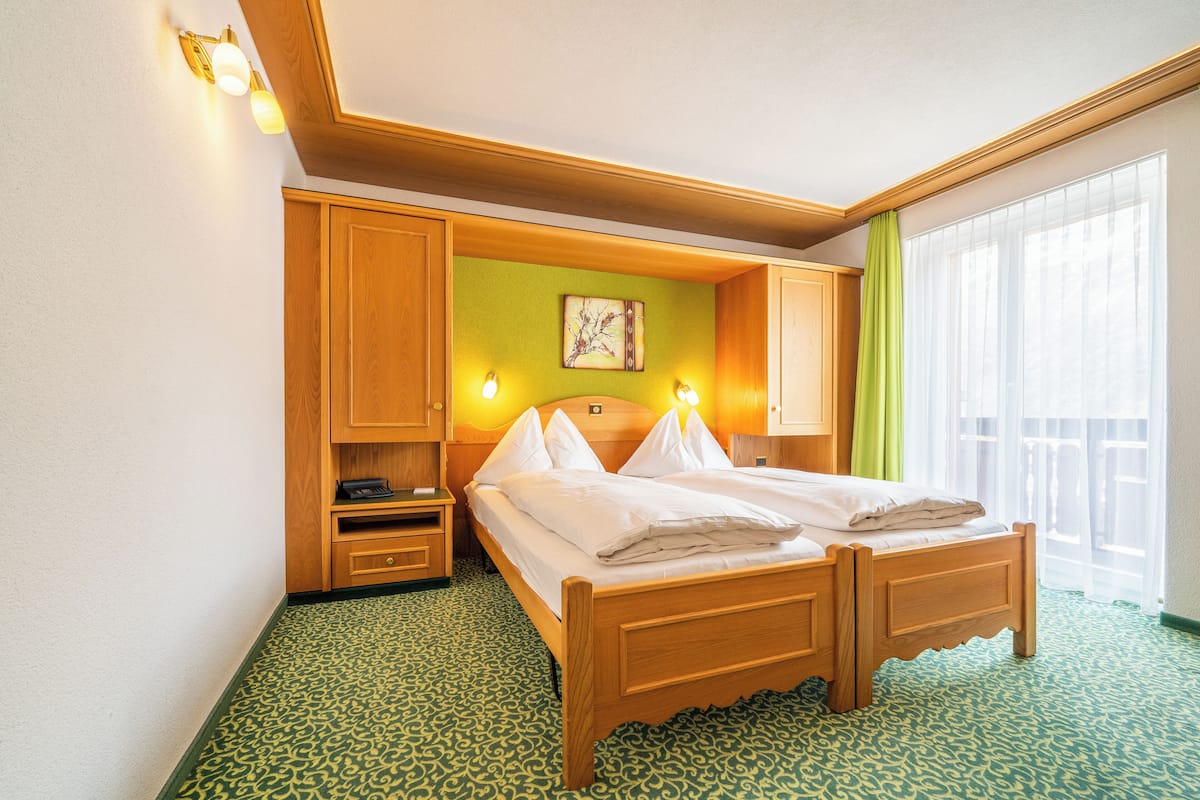 Quarto de hotel com cama de casal, cabeceira de madeira, carpete com padrão verde, guarda-roupa de madeira, abajures e uma janela com cortinas translúcidas.