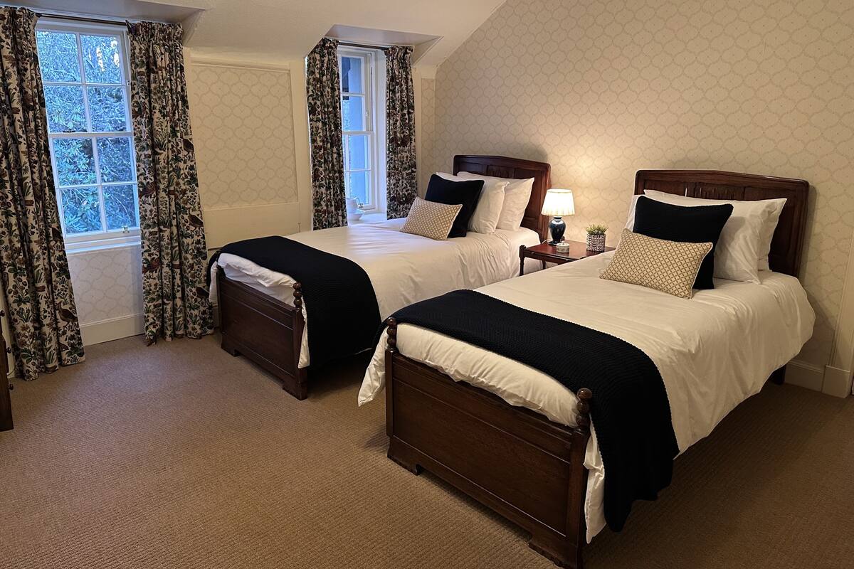 Quarto de hotel com duas camas, uma mesinha de cabeceira, um abajur e uma janela com cortinas.