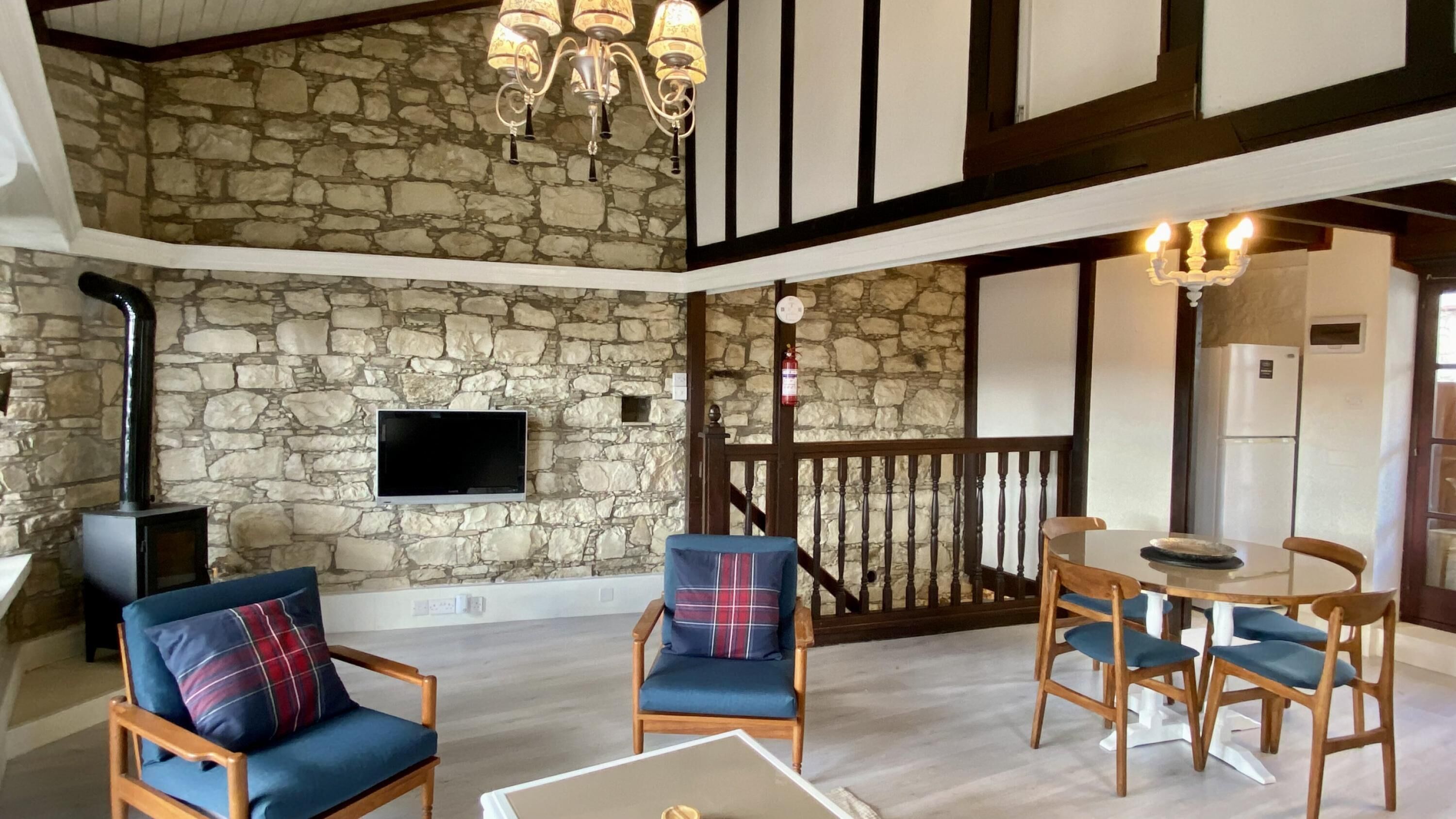 Uma sala de estar acolhedora com paredes de pedra, lareira e televisão. Há duas poltronas azuis com almofadas xadrez e uma mesa de madeira com cadeiras.