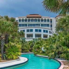 Um hotel com piscina, rodeado por palmeiras e um edifício grande com várias varandas.