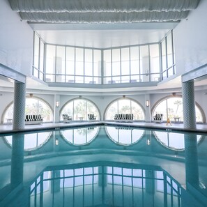 Piscina interna com vista ampla do céu através de grandes janelas, cercada por espreguiçadeiras e teto alto.