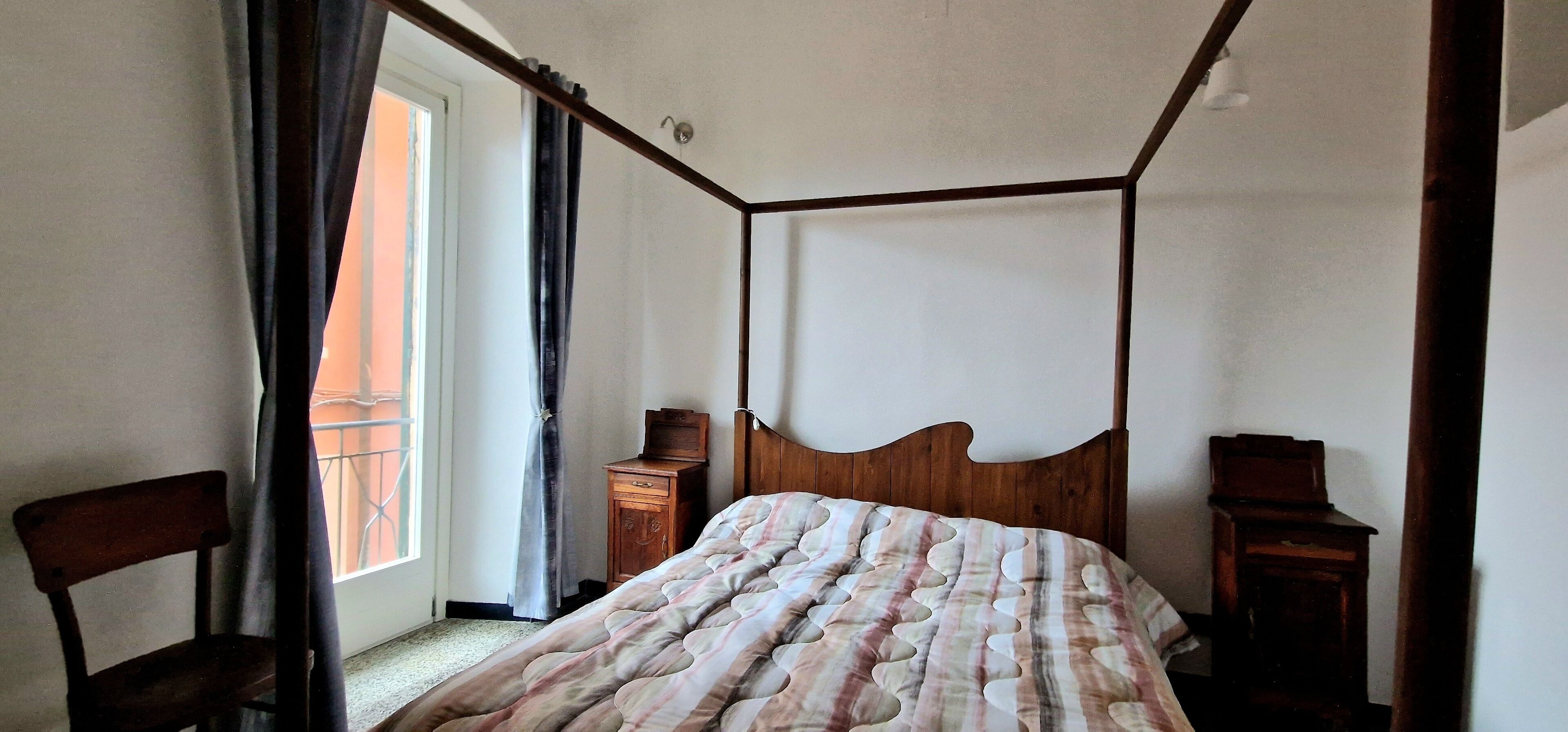 Uma cama de dossel com colcha, uma mesa de cabeceira de madeira, uma cadeira e uma janela com cortinas.