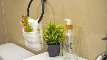 Casa de banho com um porta-toalhas com uma toalha branca e uma planta num vaso, uma garrafa de vidro transparente com dispensador e uma parede revestida a azulejos bege.