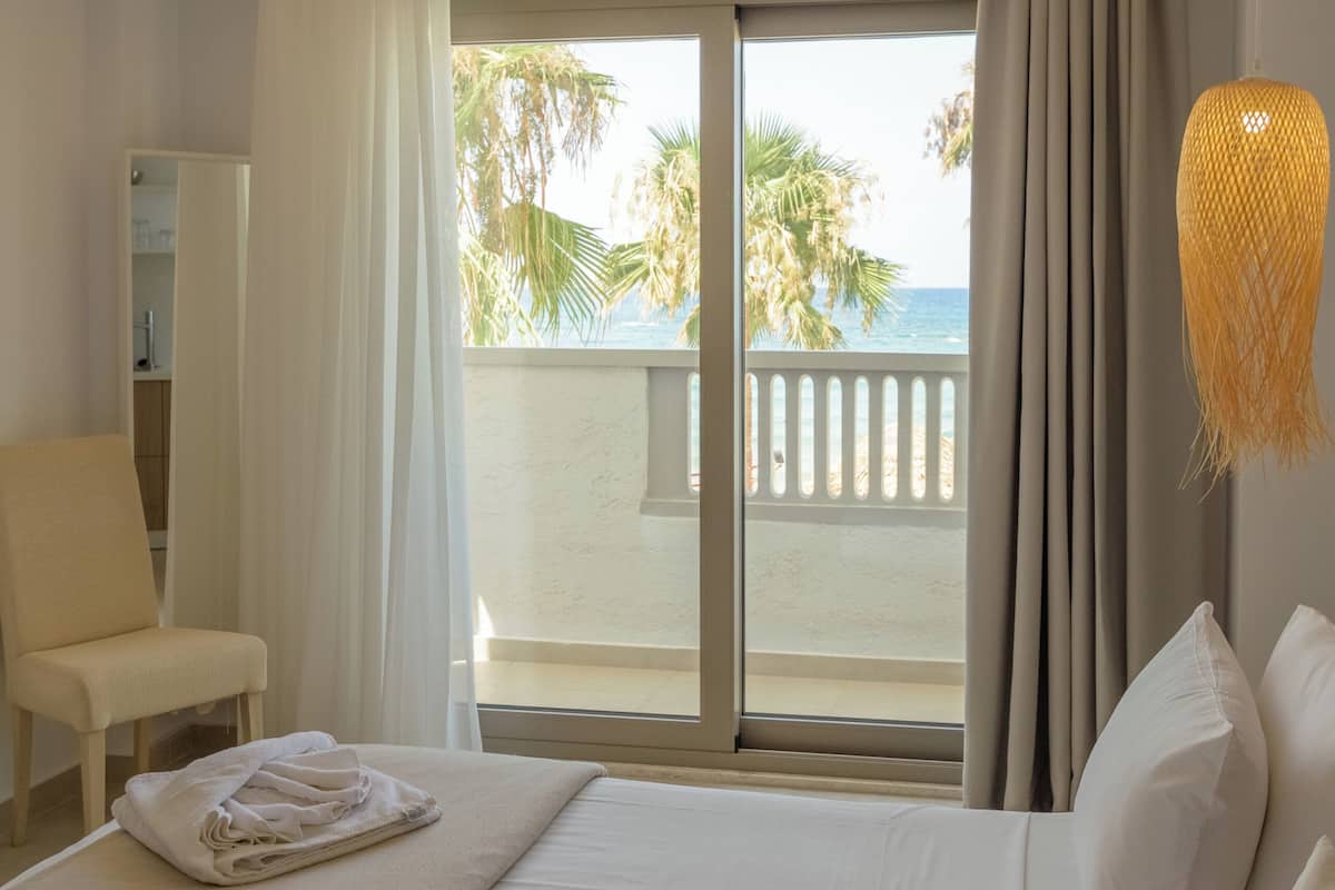Quarto de hotel com cama, cadeira, varanda com vista para a praia e palmeiras, e luminária suspensa.