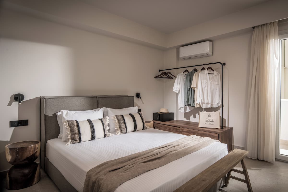 Quarto de hotel moderno com uma cama grande, mesas de cabeceira, um banco, um cabideiro com roupas e um ar condicionado fixado na parede.
