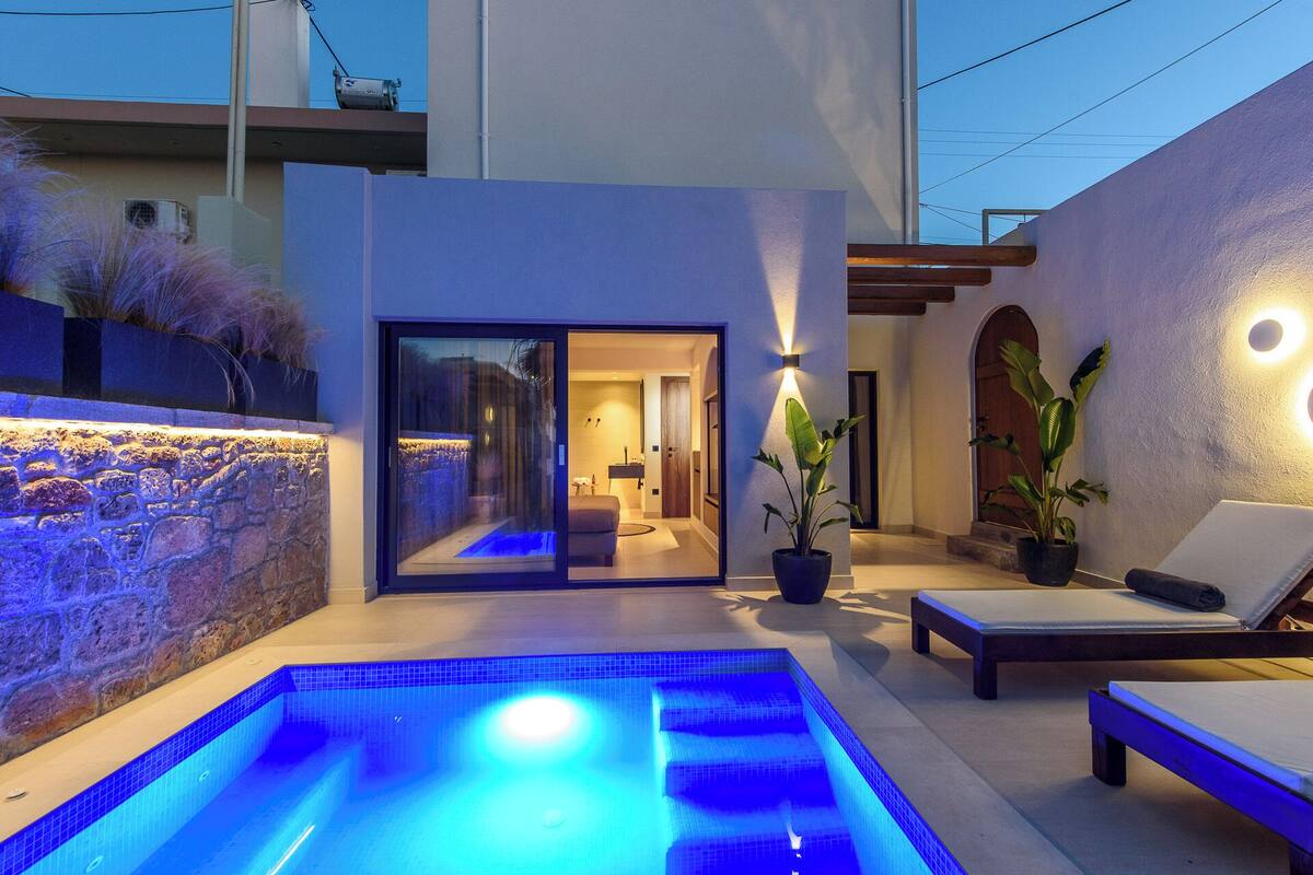 Uma área de piscina moderna ao ar livre, com porta de vidro que dá para um espaço interior bem iluminado, uma parede de pedra e espreguiçadeiras.