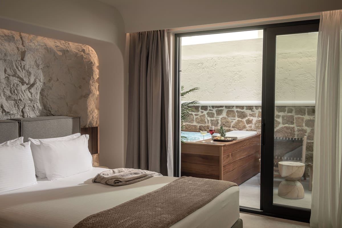 Quarto de hotel com uma cama grande, parede de pedra, escrivaninha de madeira com vista e varanda com parede de pedra e uma planta.