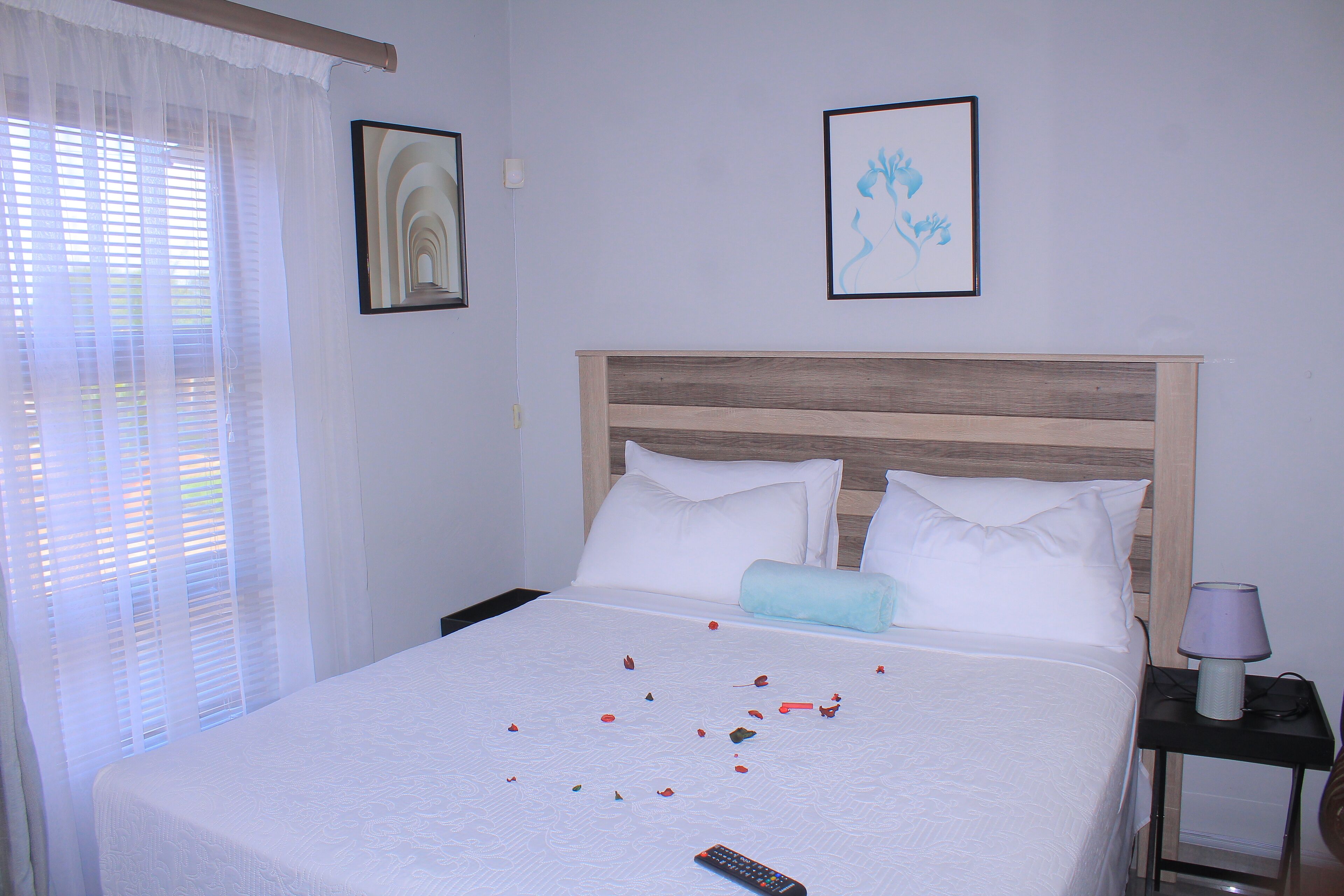 Quarto de hotel com uma cama grande, mesa de cabeceira com abajur e duas fotos emolduradas na parede.