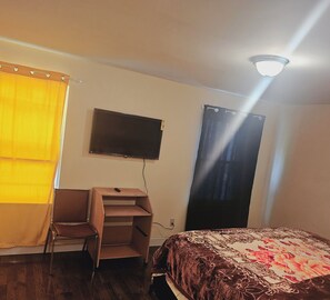 Quarto com cama, escrivaninha, cadeira, televisão na parede e janela com cortinas.