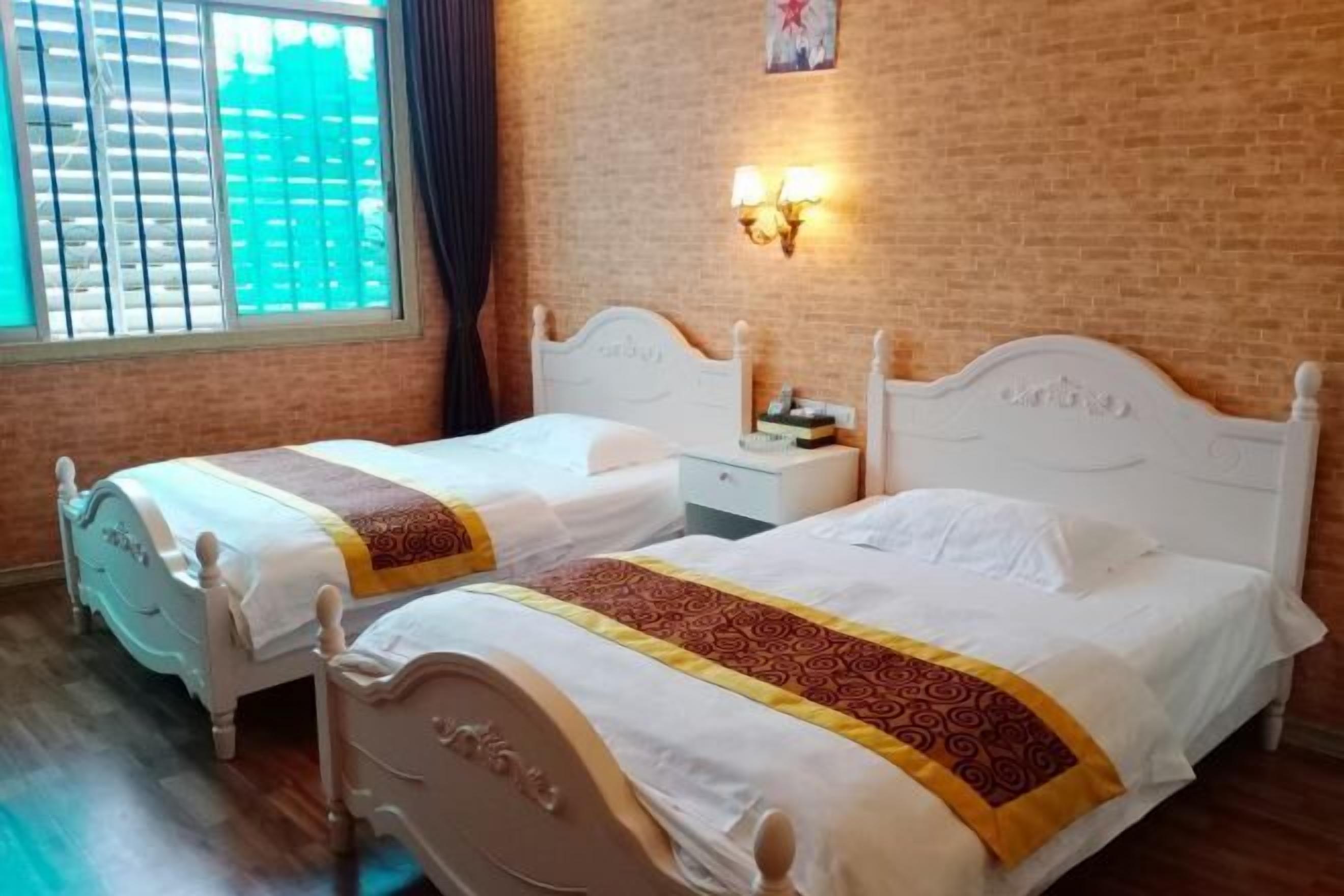 Quarto de hotel com duas camas brancas, parede de tijolos, janela com persianas e mesa de cabeceira com abajur.