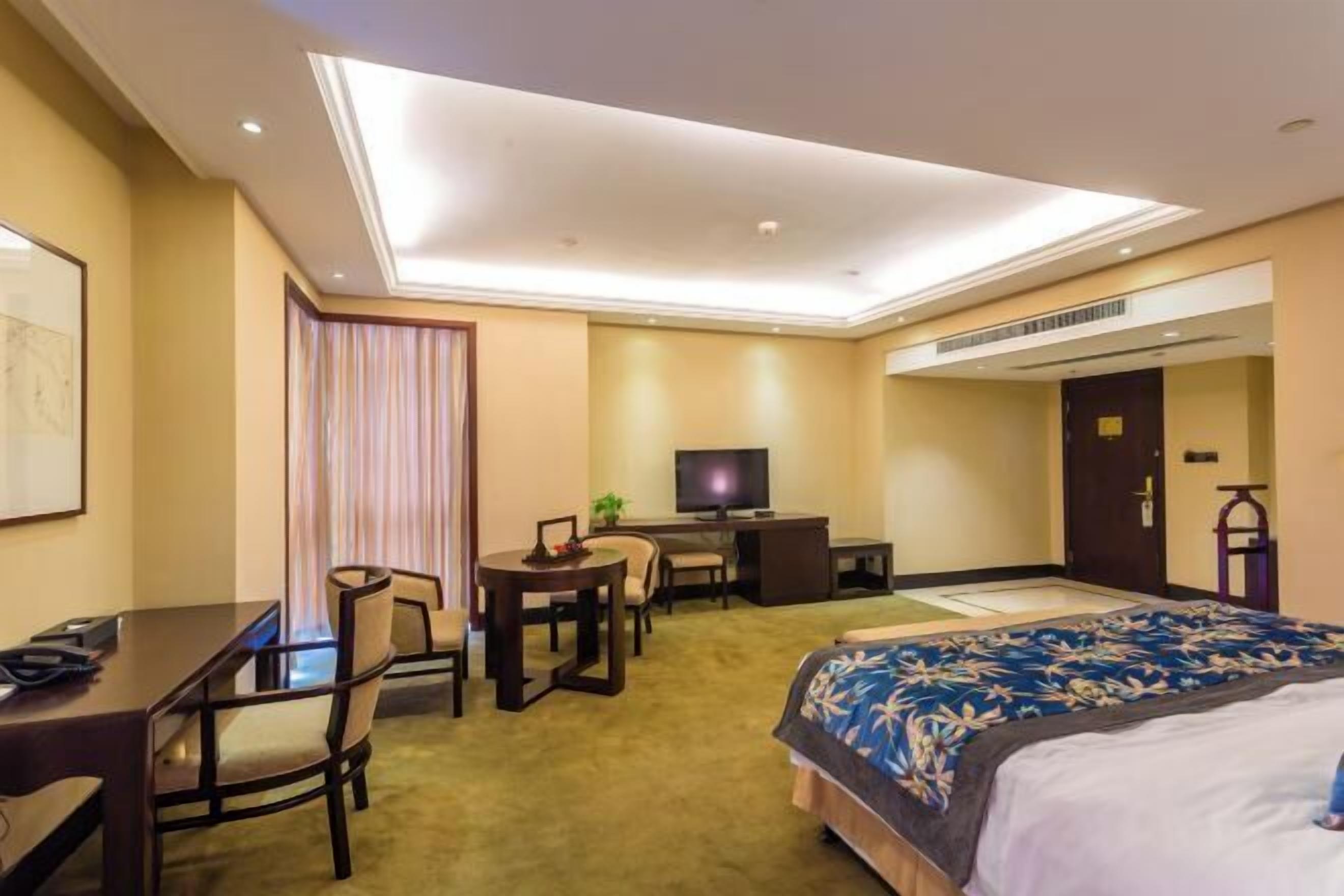Quarto de hotel com cama, escrivaninha, cadeiras, televisão e uma grande janela com cortinas.