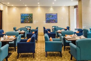 Uma área de refeições de hotel com fileiras de cadeiras e mesas azuis-esverdeadas, obras de arte nas paredes e piso estampado.