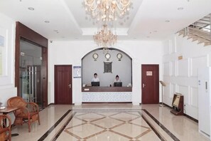 Um saguão de hotel com balcão de recepção, dois recepcionistas, um candelabro, balcão de check-in e piso ladrilhado.