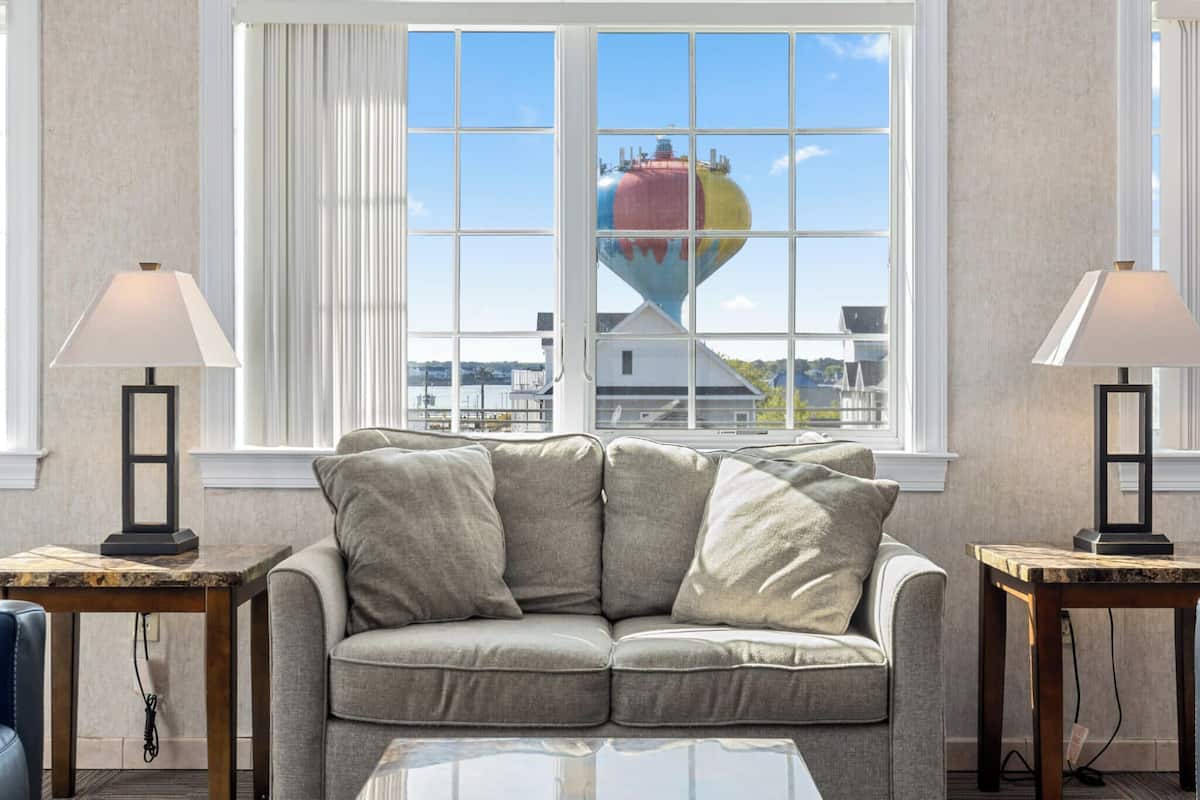 Sala de estar com sofá, duas luminárias, uma mesa e janela com vista para uma torre d'água colorida.