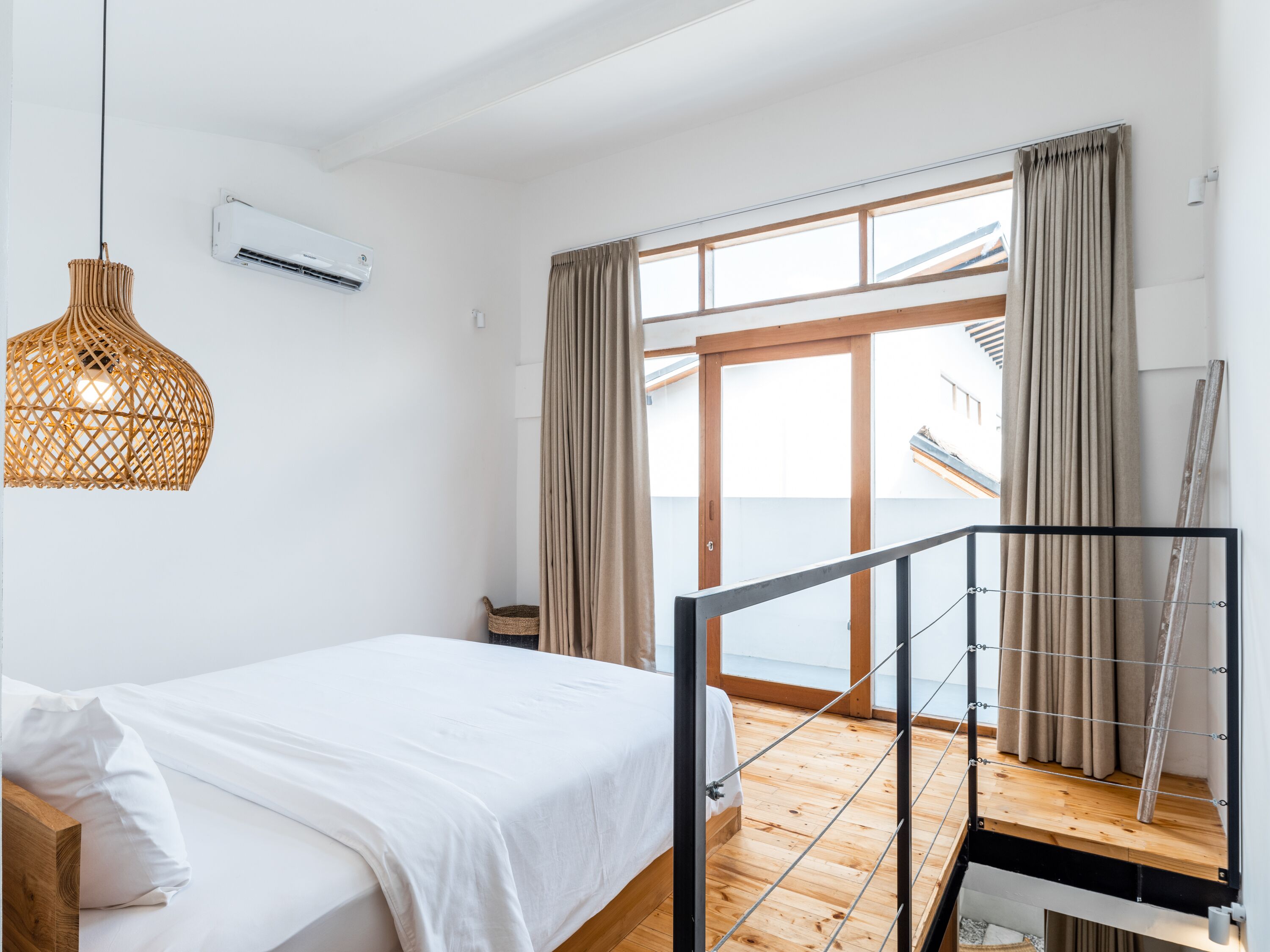 Quarto com cama, luminária pendente, ar condicionado, janela com cortinas e vista para o exterior.