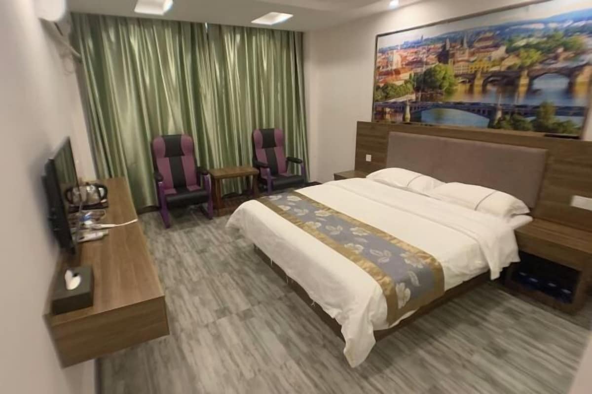 Quarto de hotel com uma cama, duas cadeiras, uma mesinha, uma televisão e um quadro na parede.