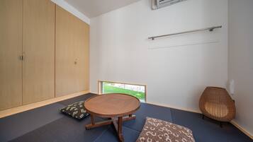 Quarto com chão de esteira de tatame, mesa de madeira redonda, armário de madeira baixo, cadeira de vime e vista para um jardim pela janela.