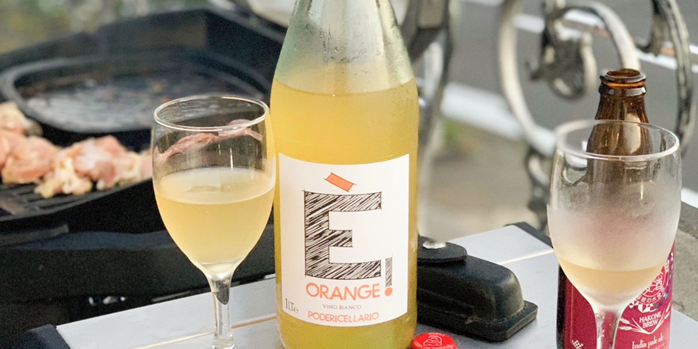 Uma garrafa de vinho laranja e dois copos sobre uma mesa, com uma churrasqueira ao fundo.