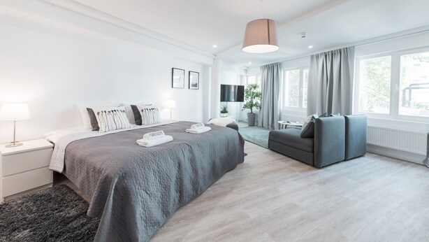 Quarto moderno com uma cama grande, um sofá cinza e vista para o exterior através de amplas janelas.