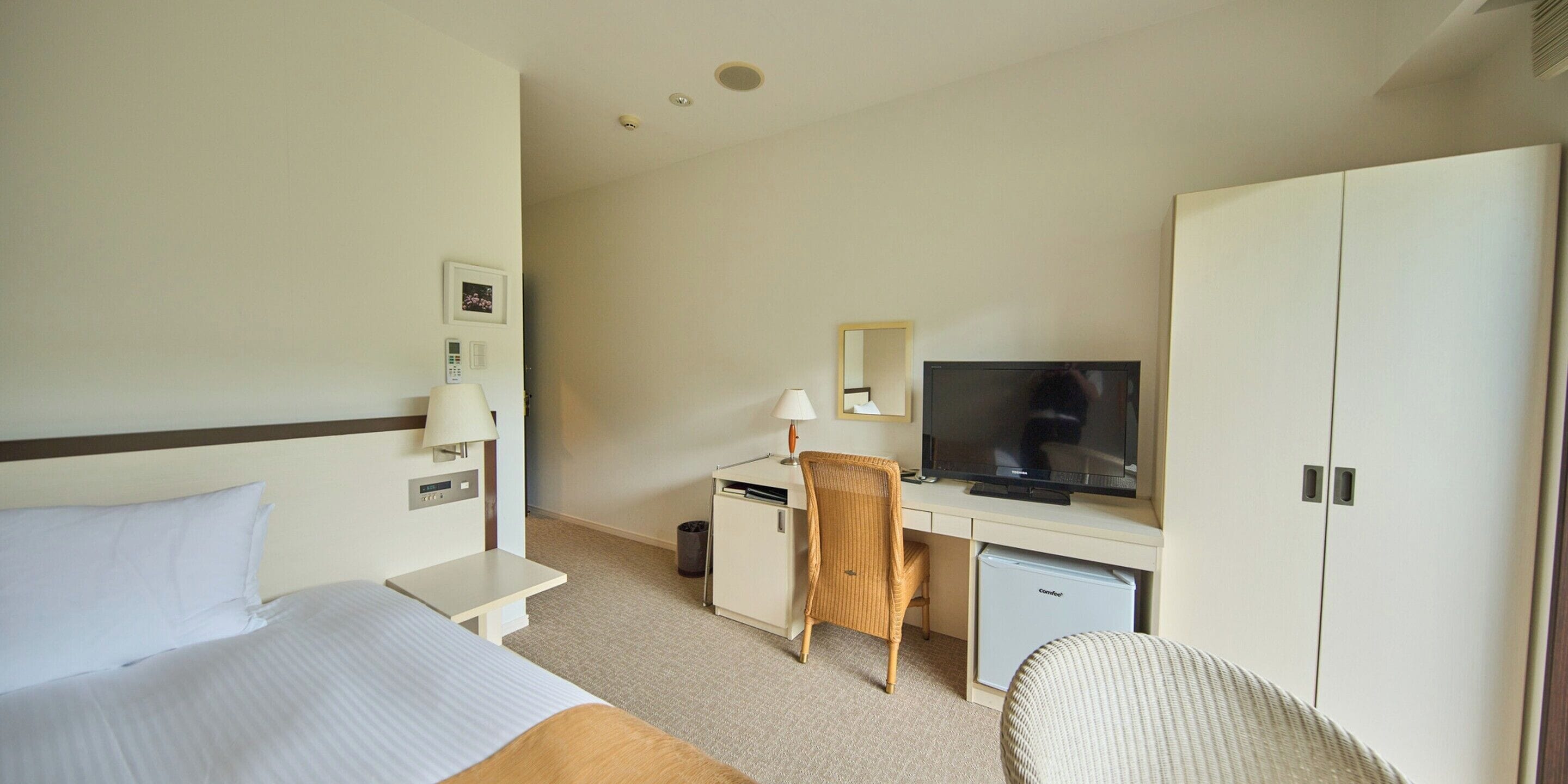 Quarto de hotel com cama, escrivaninha, cadeira, televisão e guarda-roupa.
